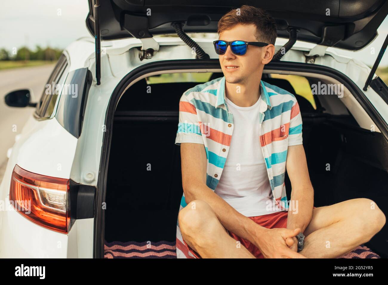 Young man in sunglasses, sitting in the trunk of a car, outdoors on the ...