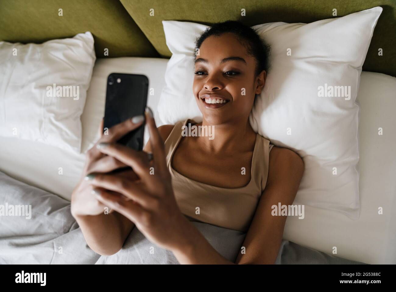 Happy african american girl using mobile phone while lying in bed after ...