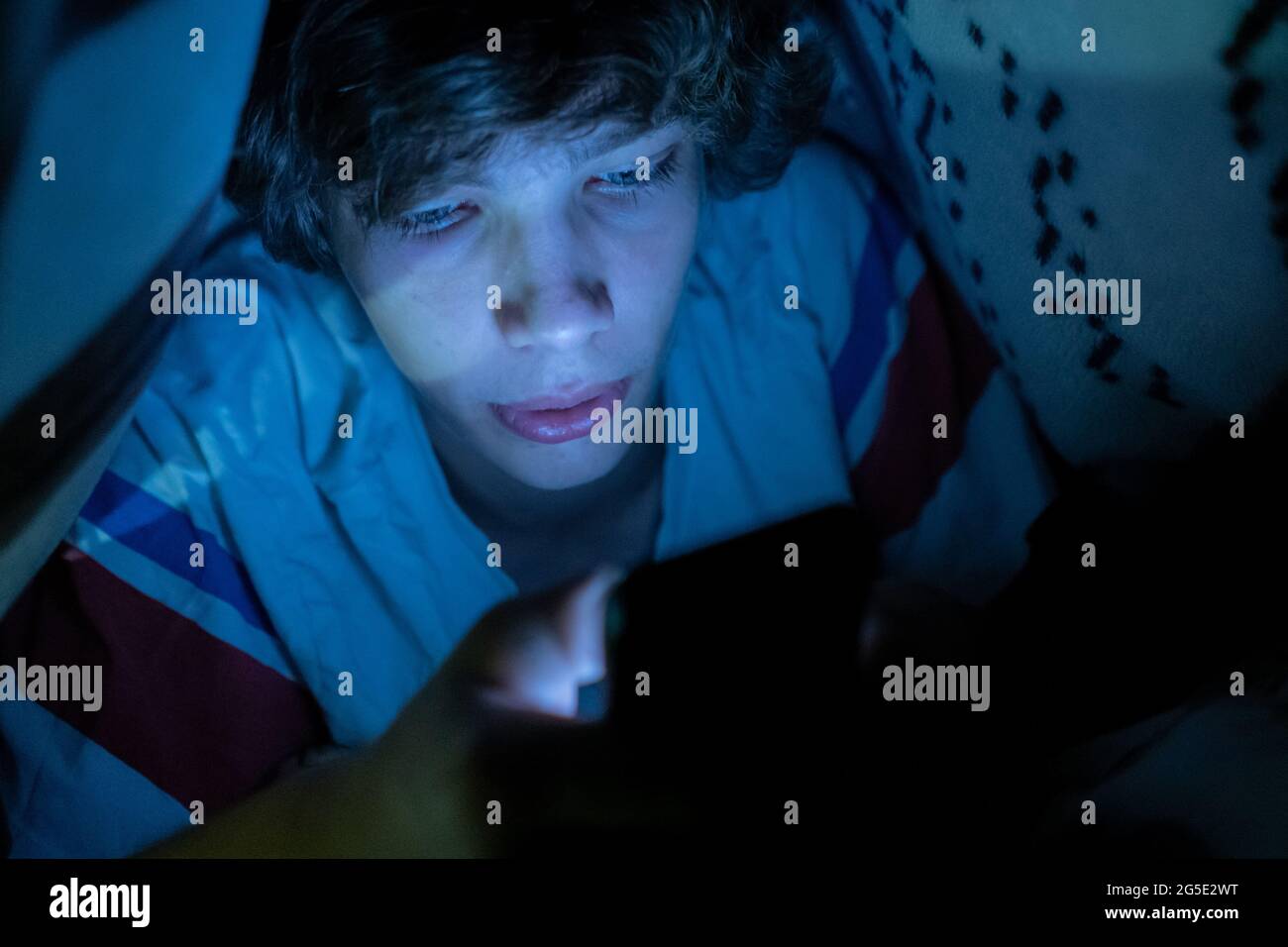 young boy using a smartphone lying in bed under a blanket at night ...