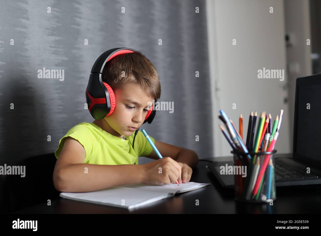 Boy listening to school lessons on a laptop with headset and writing in ...