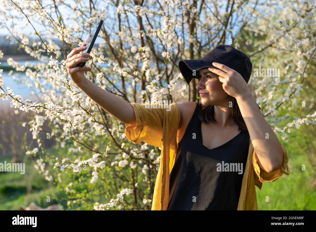 A stylish girl in a hat makes a selfie at sunset near flowering trees ...