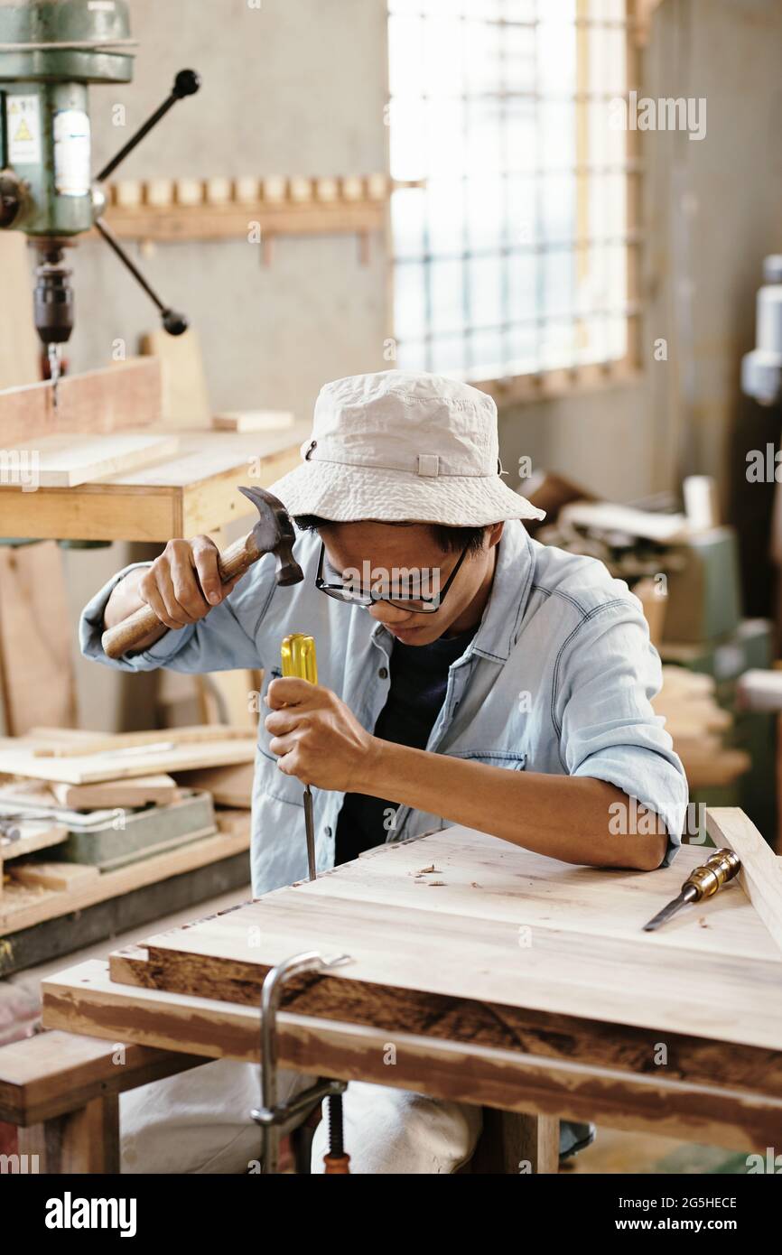 Serious young carpenter working in workshop and carving wood with ...