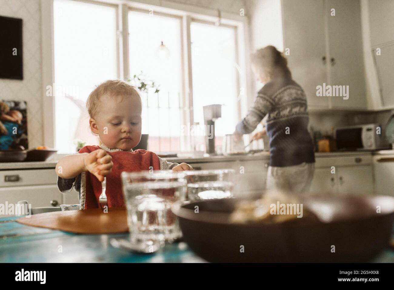 Girl sitting at dining table while mother prepares food in kitchen ...