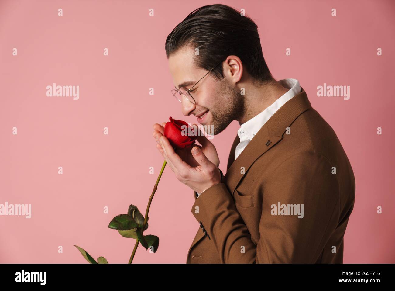 Happy mid aged brunette man in jacket smelling red rose over pink wall ...