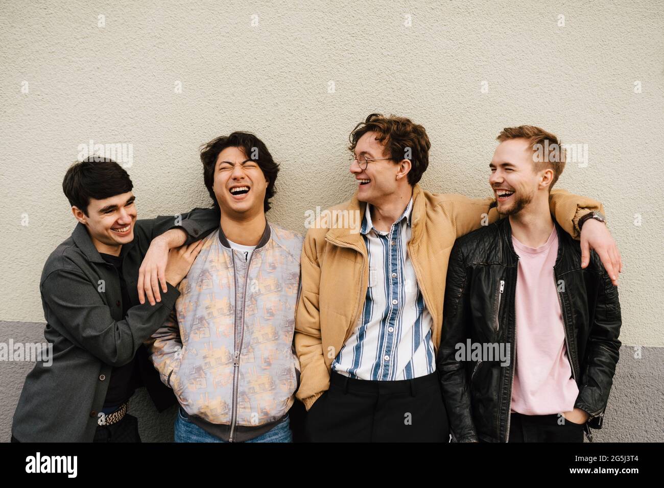 Cheerful male friends laughing while standing against beige wall Stock ...