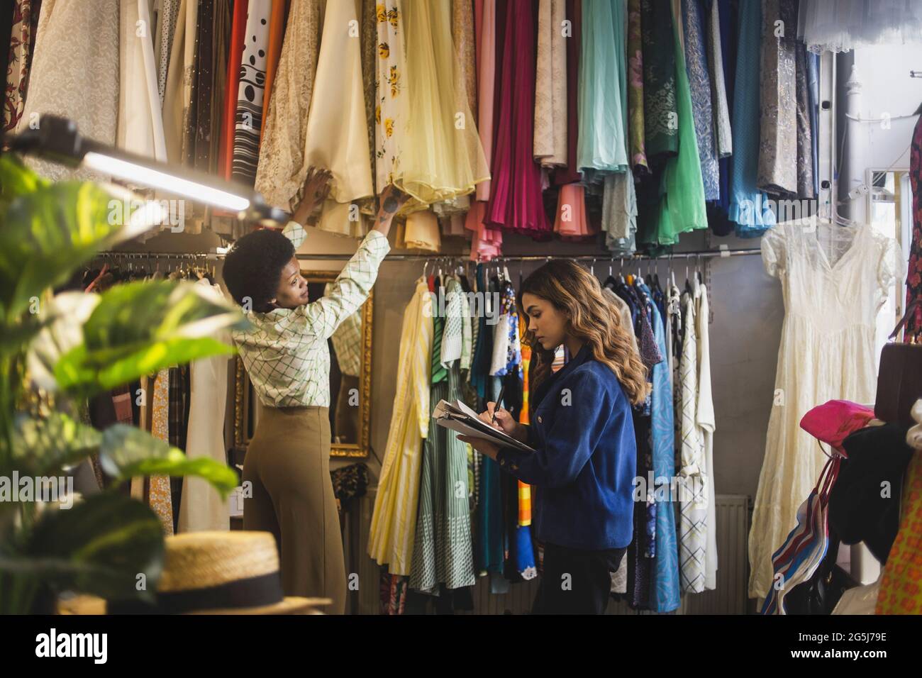 Female owner making notes while colleague arranging clothes in boutique ...