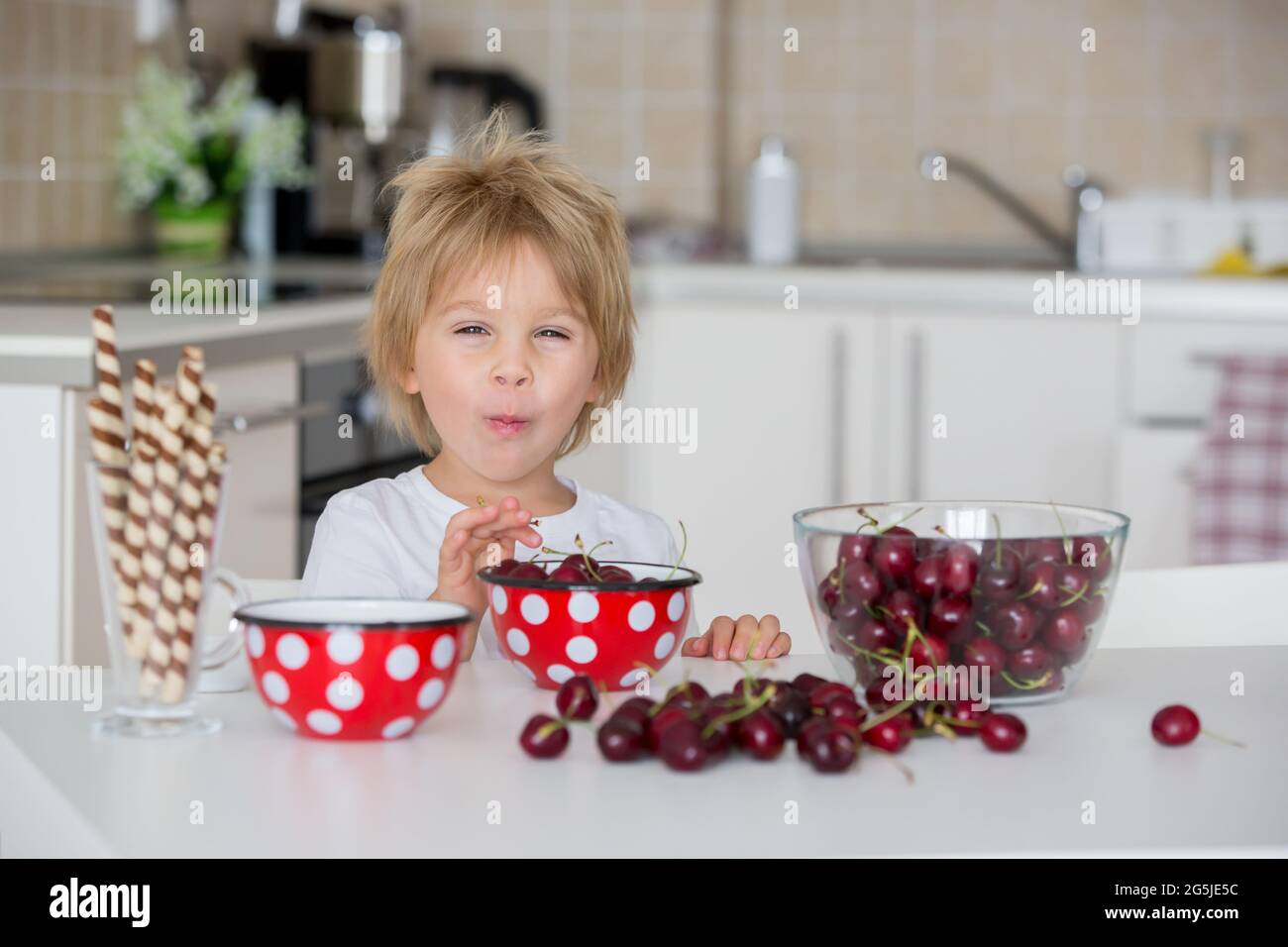 Cute blond child, toddler boy, eating cherries at home, having fun ...