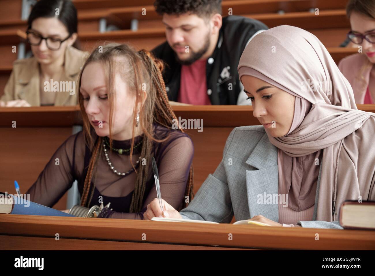 Two girls writing down lecture notes at lesson while sitting by desk ...