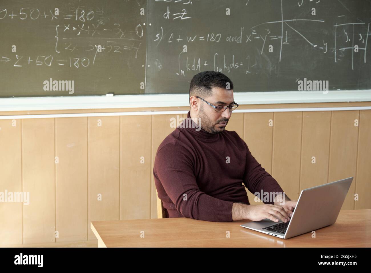 Serious teacher typing by desk against blackboard in classroom Stock ...