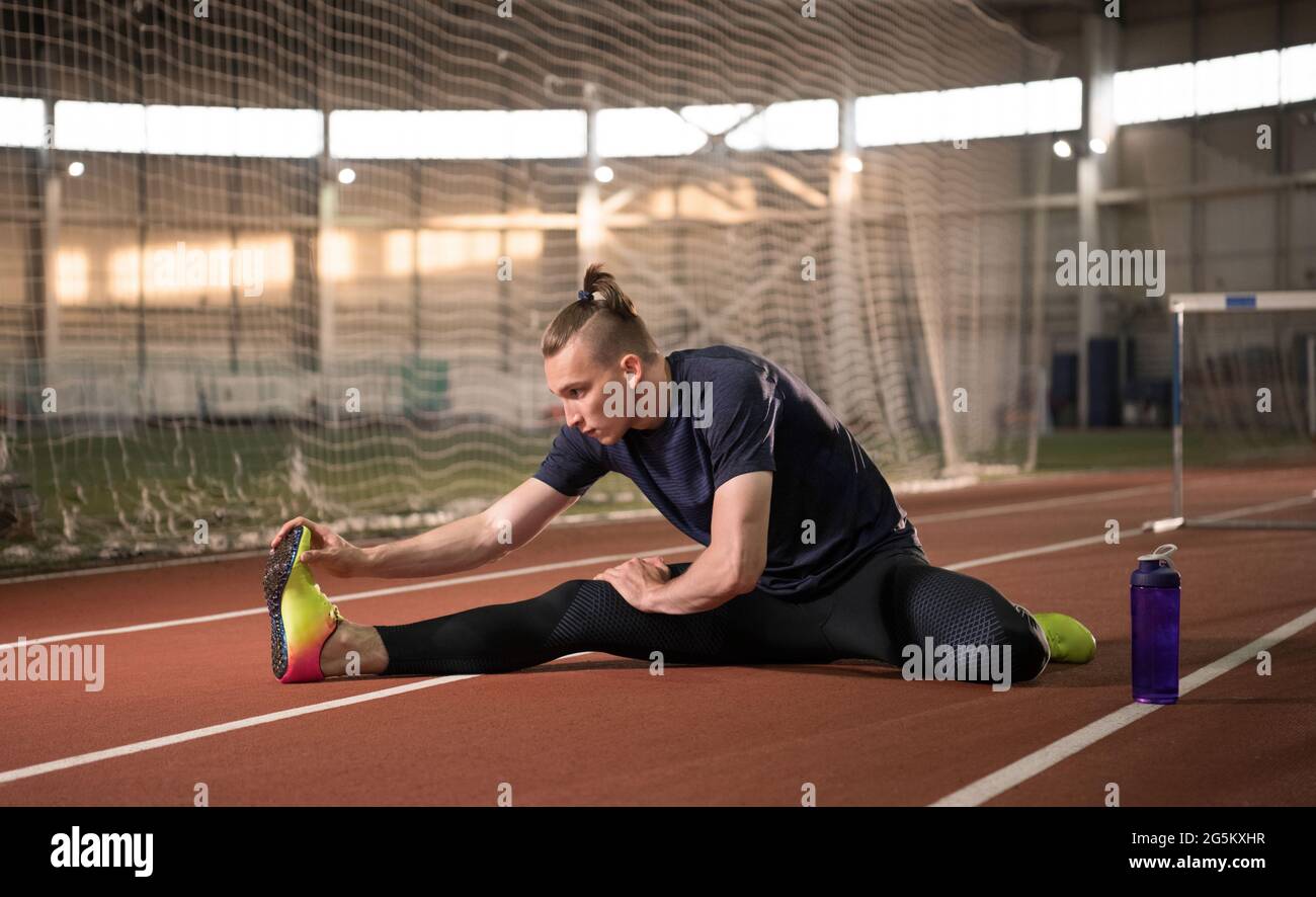 Young sportsman bending to leg and stretching while sitting on track on ...