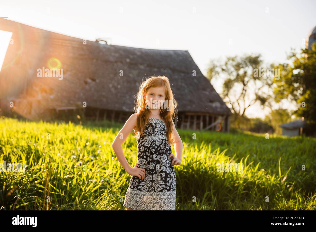 Young girl with red hair standing in tall grass, barn in background ...