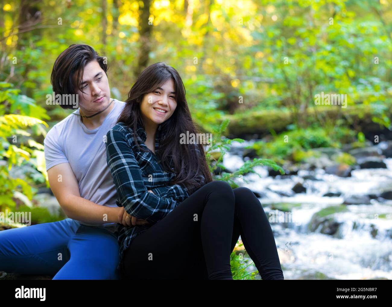 Biracial young couple sitting along rocky stream in green woods hugging ...