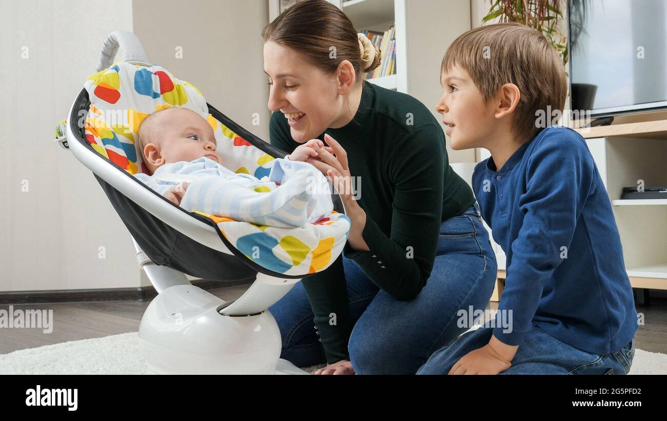 Cute smiling baby boy rocking in chair and looking at mother ond older ...