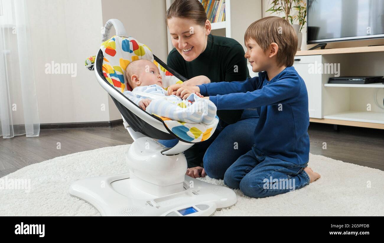 Older brother and smiling mother playing with little baby boy rocking ...