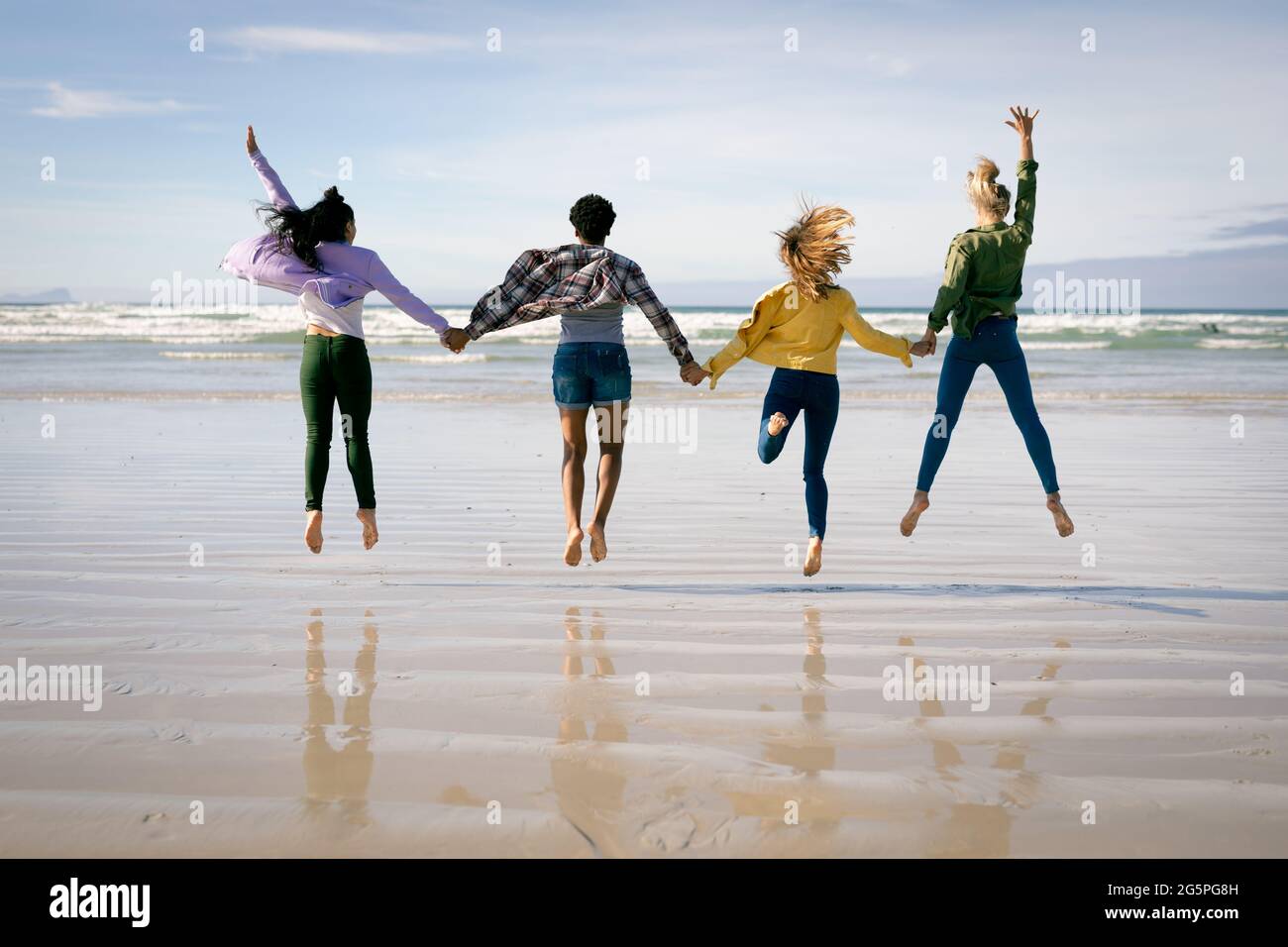 Happy group of diverse female friends having fun, walking along beach holding hands and jumping ...