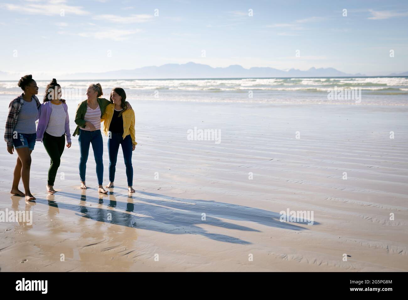 Happy group of diverse female friends having fun, walking along beach holding hands and laughing ...