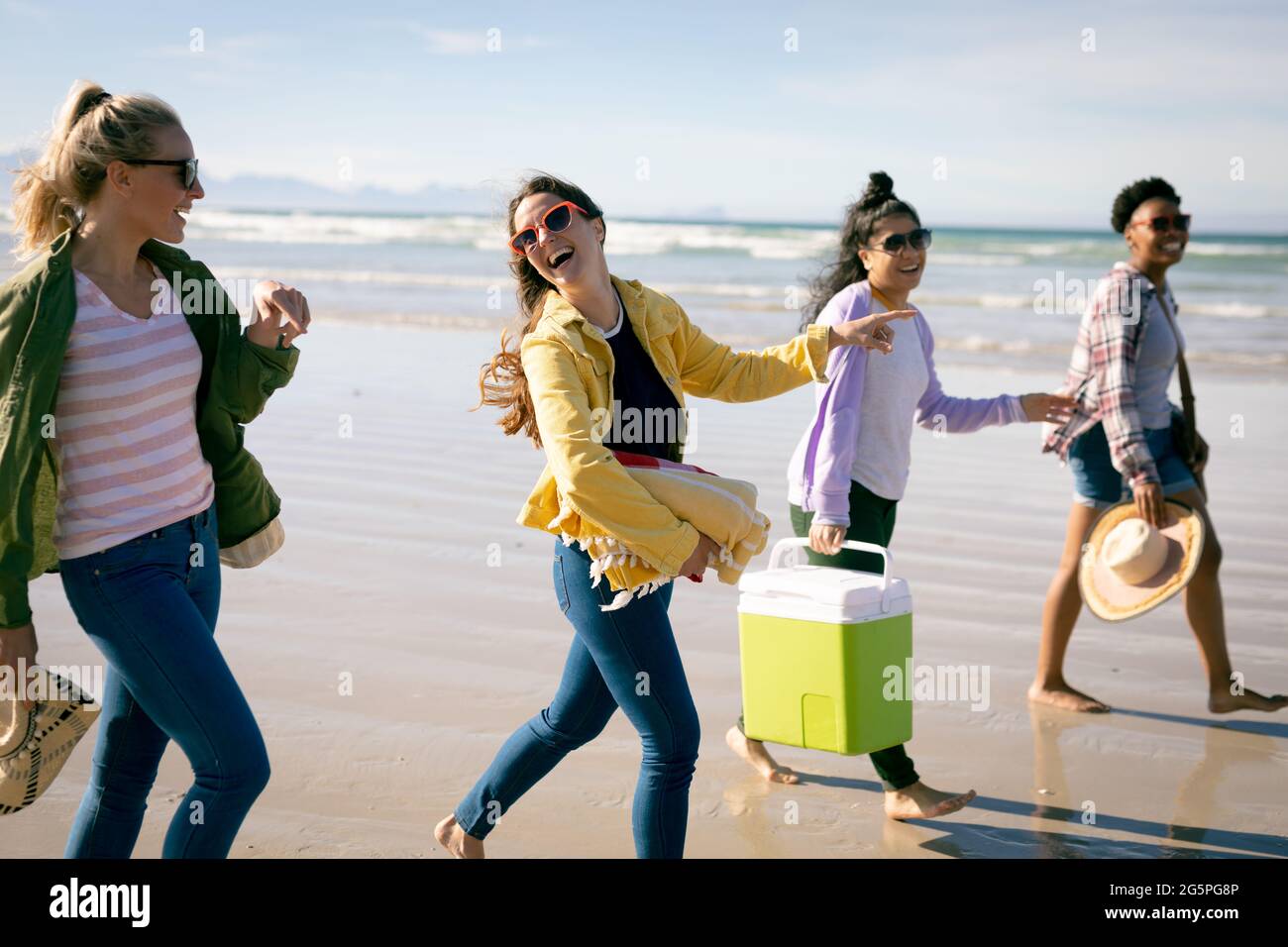 Happy group of diverse female friends having fun, walking along beach holding hands and laughing ...