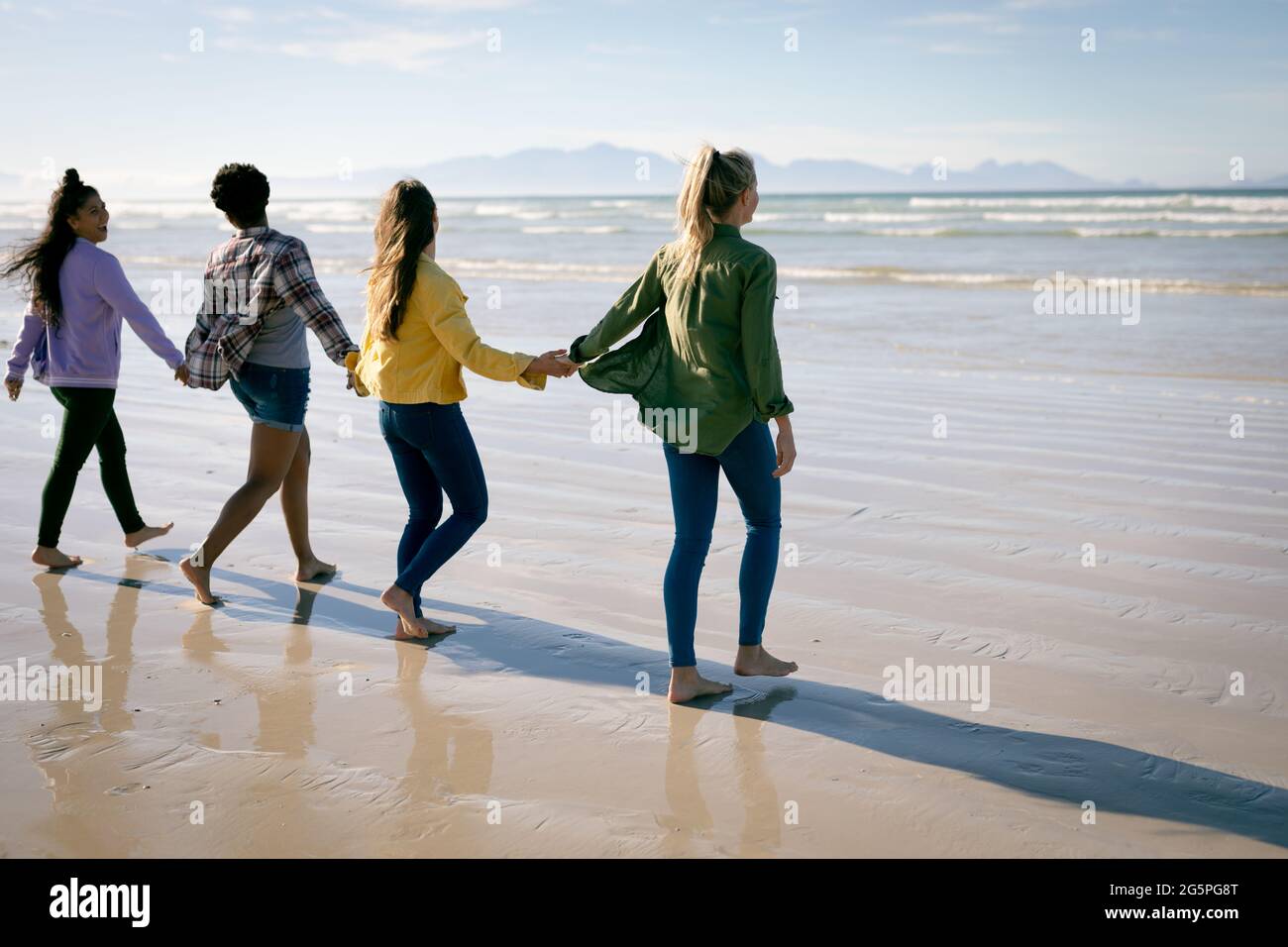 Happy group of diverse female friends having fun, walking along beach holding hands and laughing ...