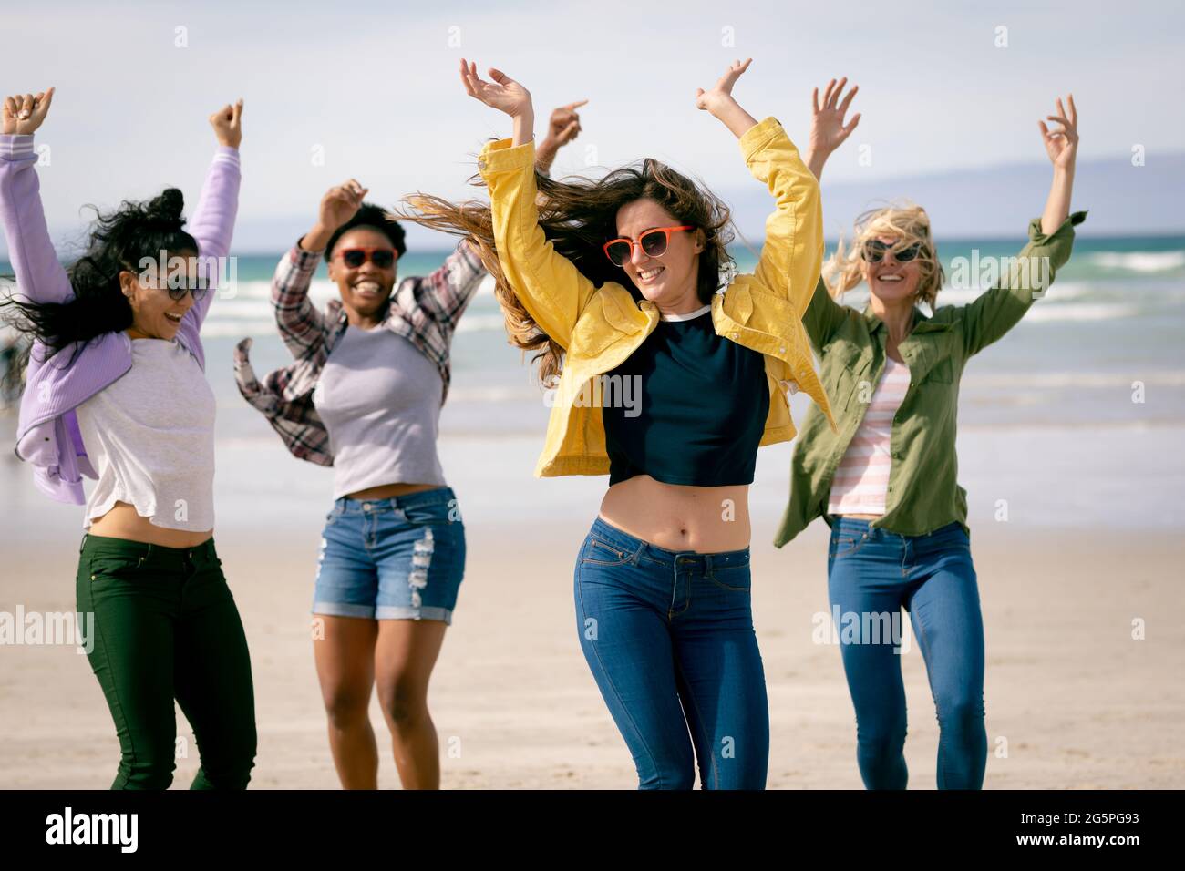 Happy group of diverse female friends having fun, walking along beach holding hands and laughing ...