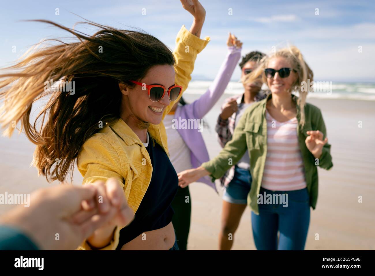 Happy group of diverse female friends having fun, walking along beach holding hands and laughing ...