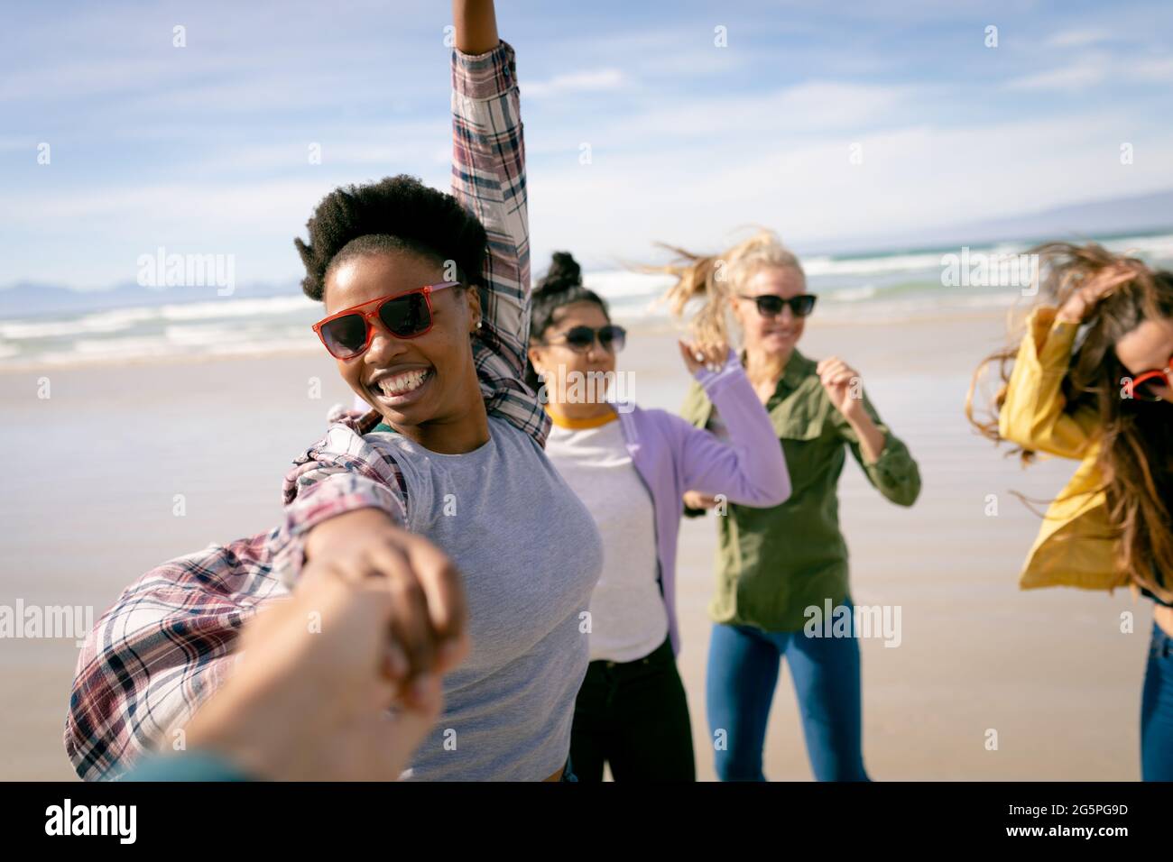 Happy group of diverse female friends having fun, walking along beach holding hands and laughing ...