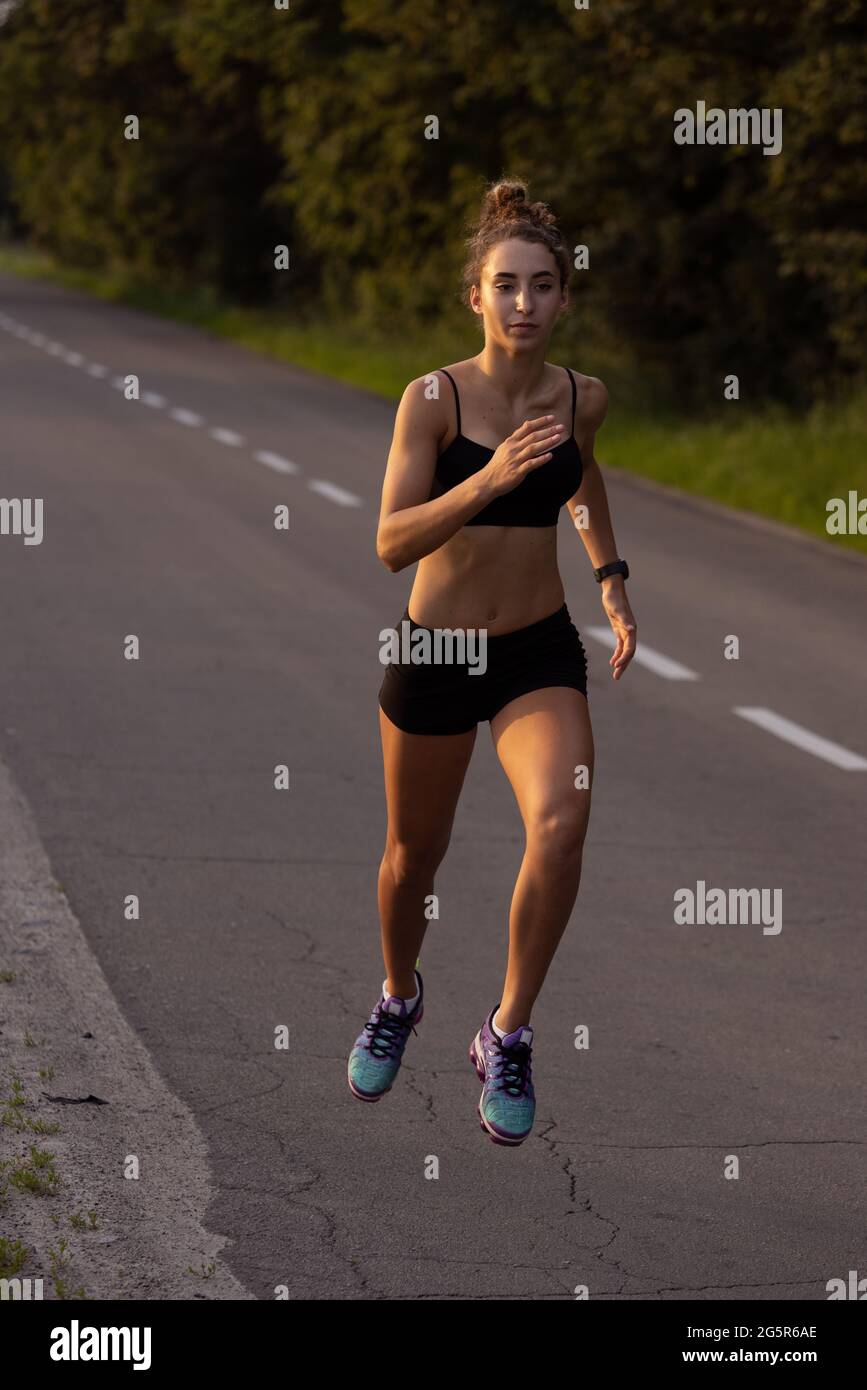 Young female runner, athlete is jogging at road in summer sunshine. Beautiful caucasian woman ...