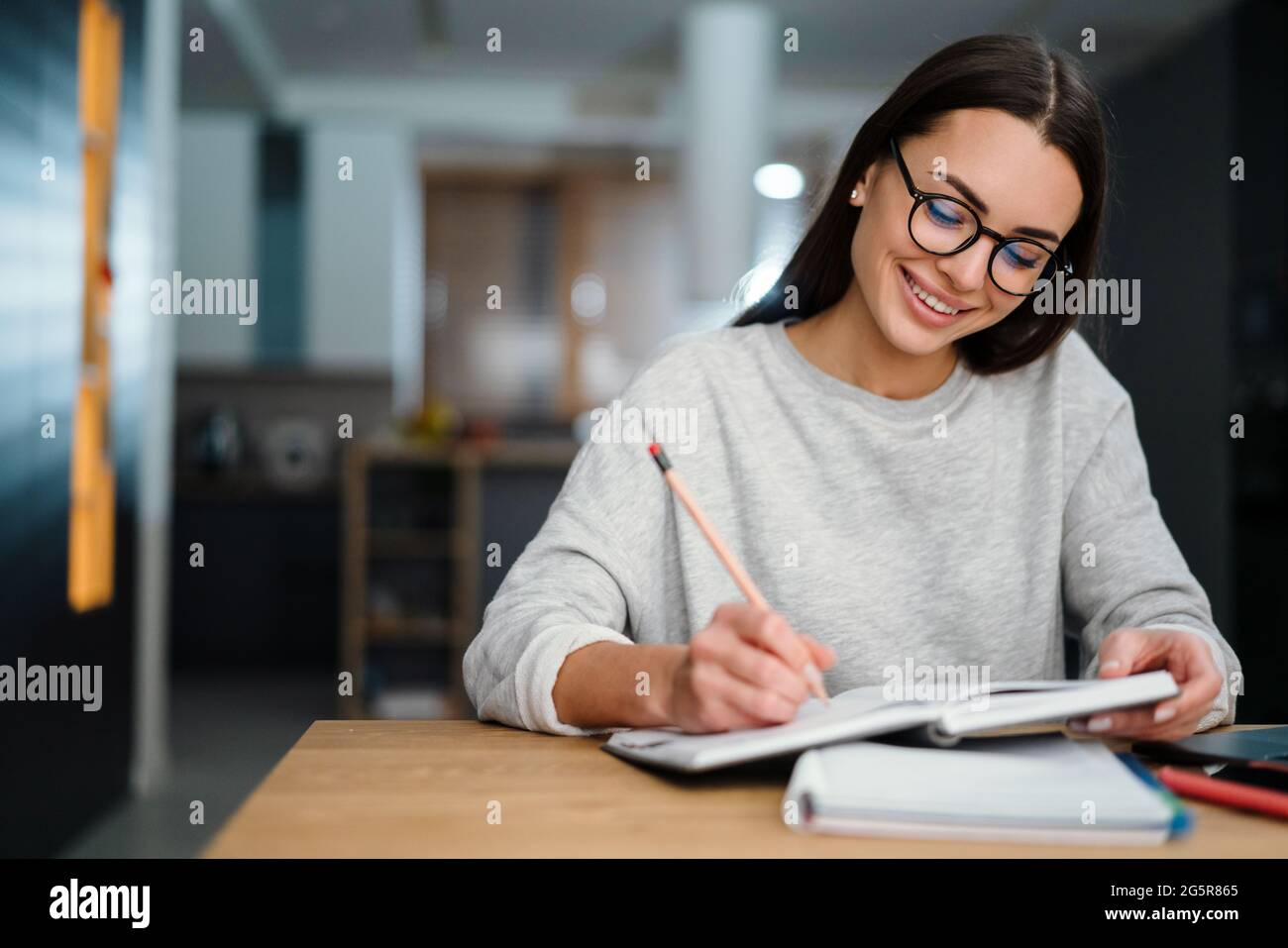 Happy young woman in eyeglasses writing down notes while working at ...