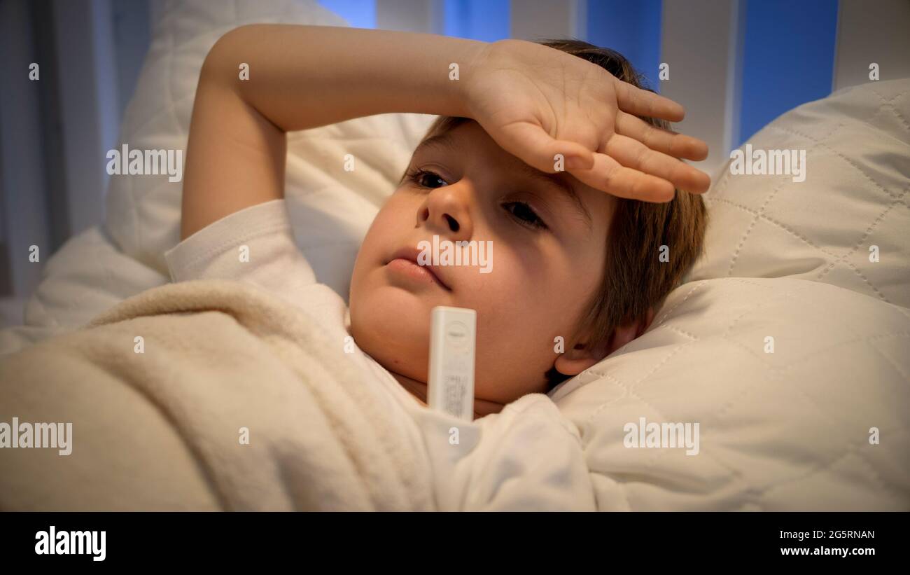 Little sick boy feeling unwell lying in bed and measuring temperature ...