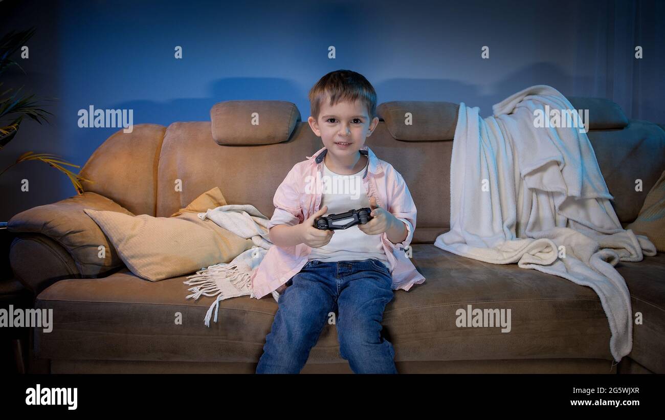 Funny excited boy playing video games on TV console on sofa at night ...