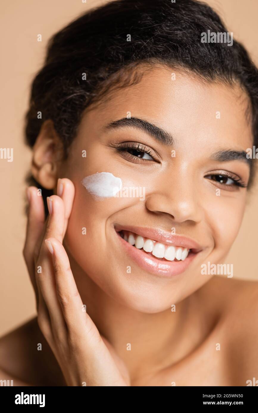 smiling african american woman applying face cream on cheek isolated on ...