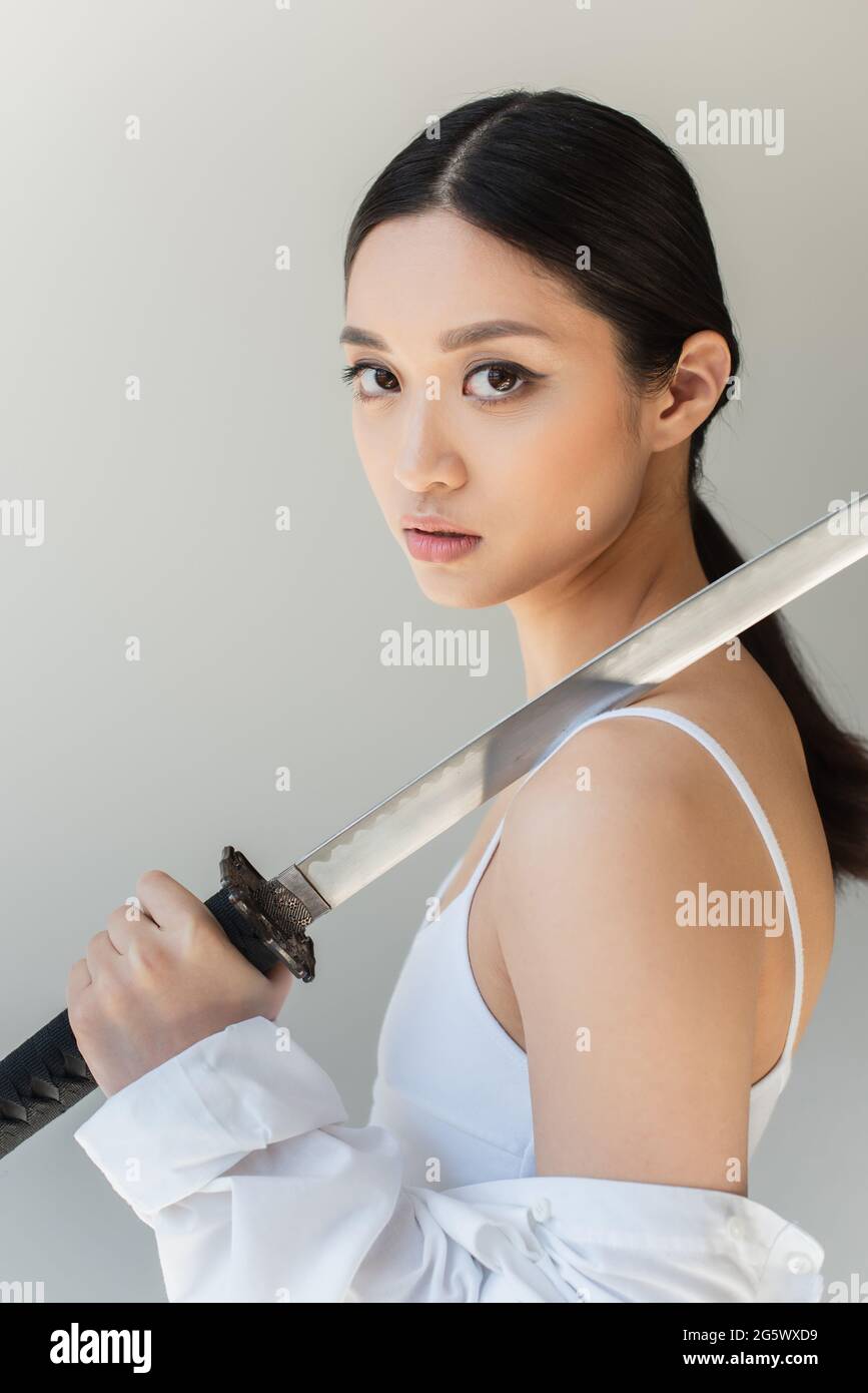 Asian woman in white clothes holding sword on shoulder isolated on grey ...