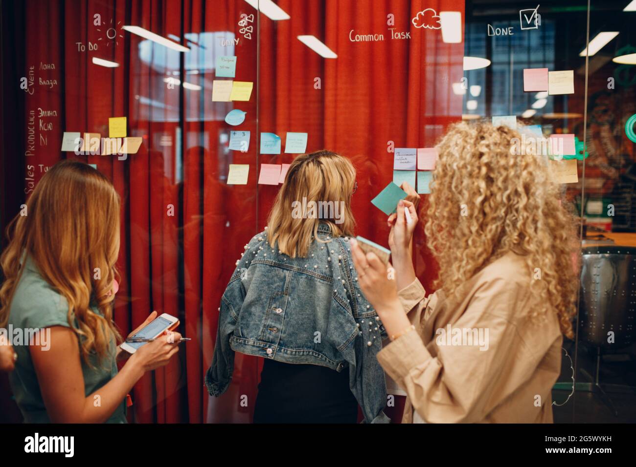 Business women writing on sticky note on glass wall with coworkers ...