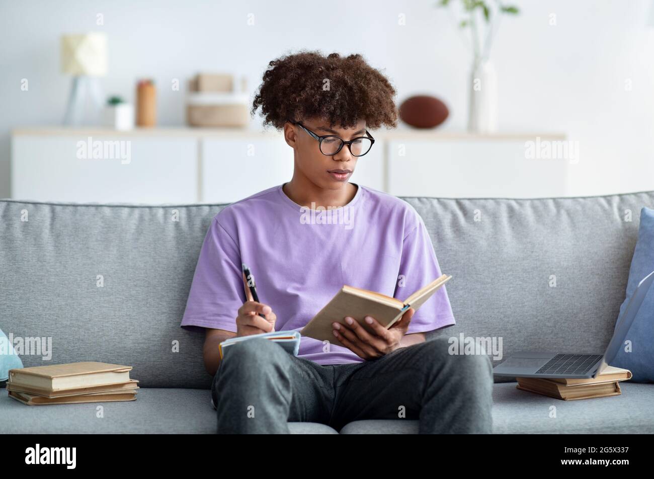 Focused black adolescent with textbook taking notes, studying at home ...