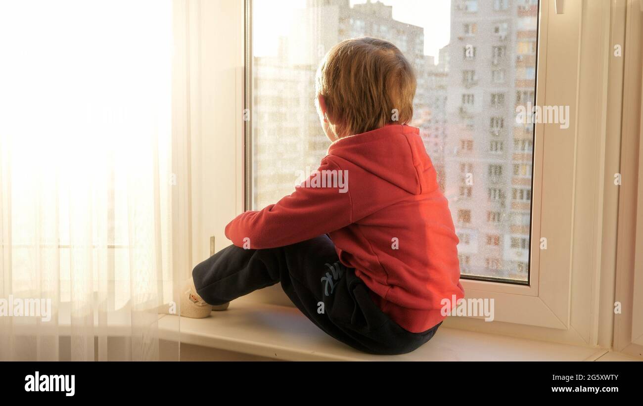 Little boy sitting on windowsill and looking on the street and sunset ...
