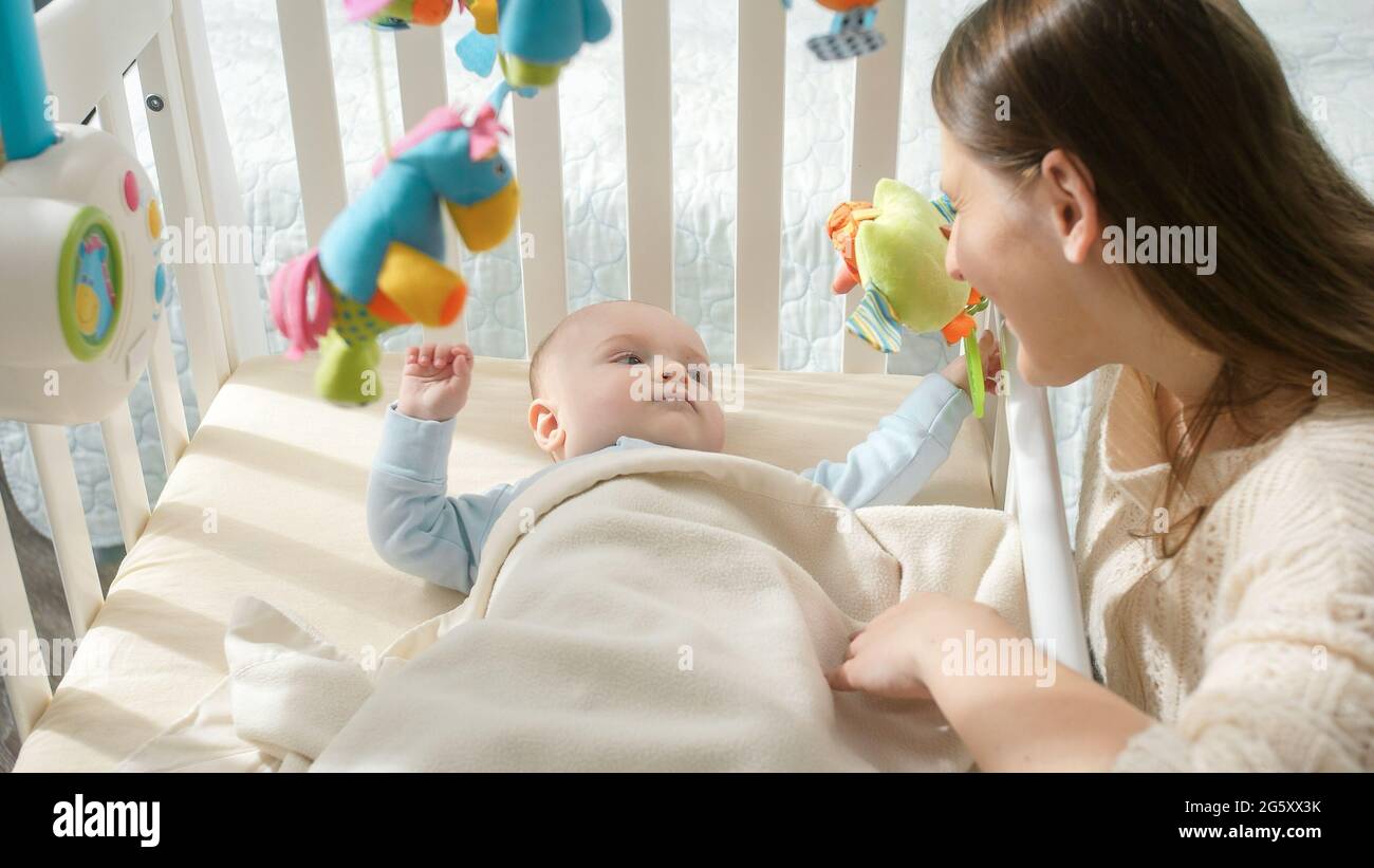 Happy smiling mother sitting at cradle and looking on her little baby ...