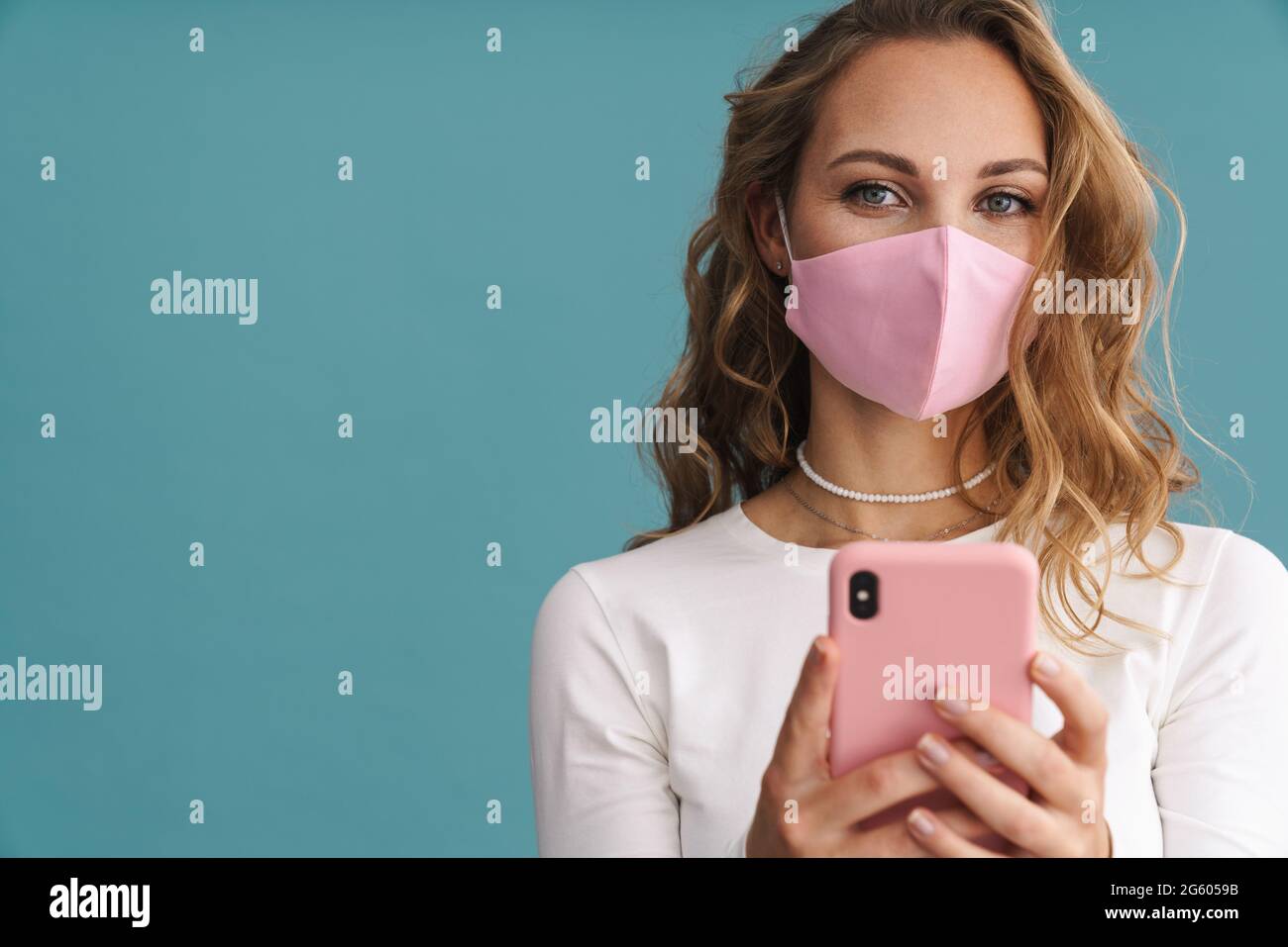 Blonde young woman wearing face mask holding mobile phone over blue ...