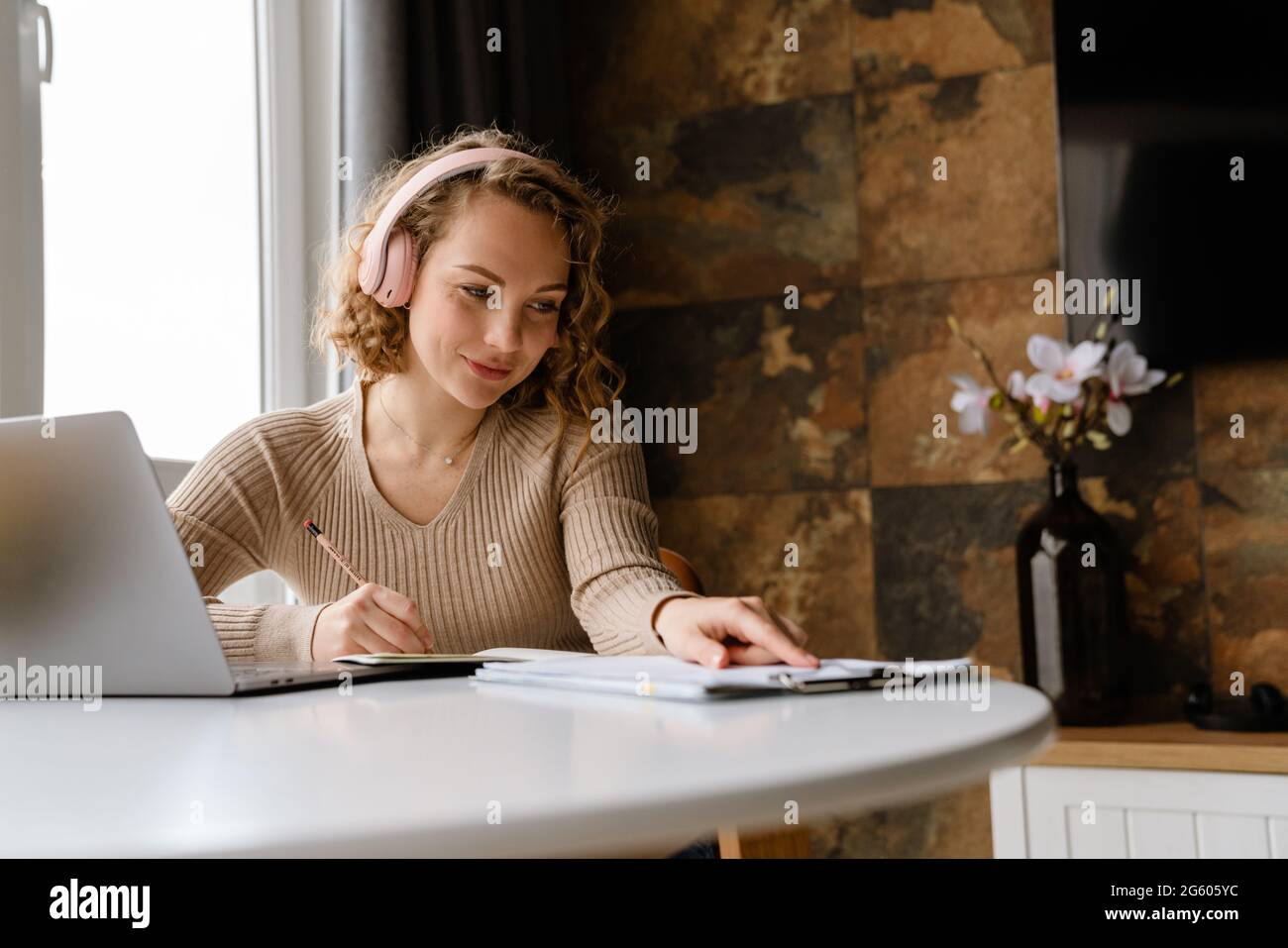 Young woman in headphones writing down notes while working with laptop ...