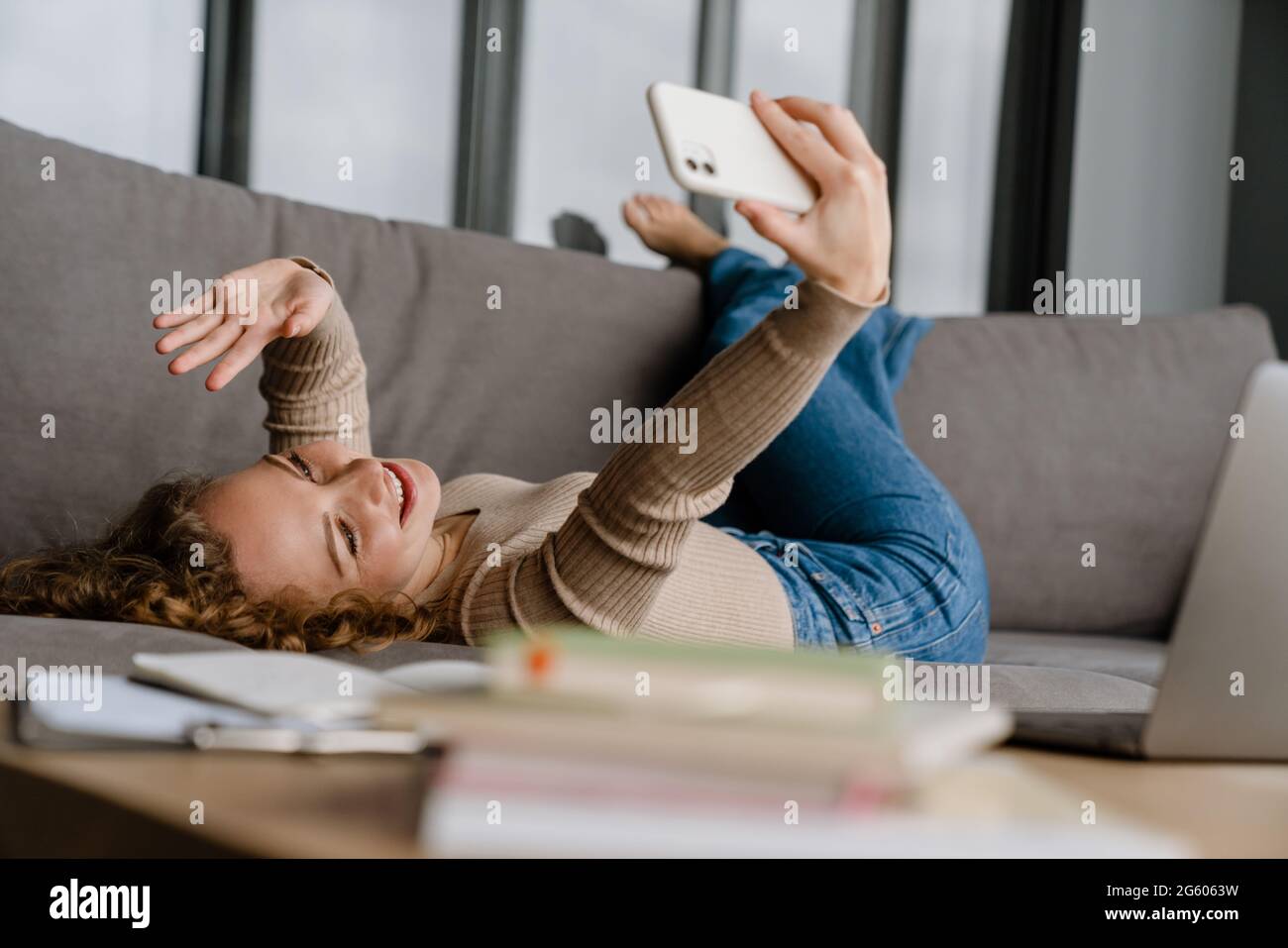 Young woman in earphone gesturing and using cellphone while lying on ...