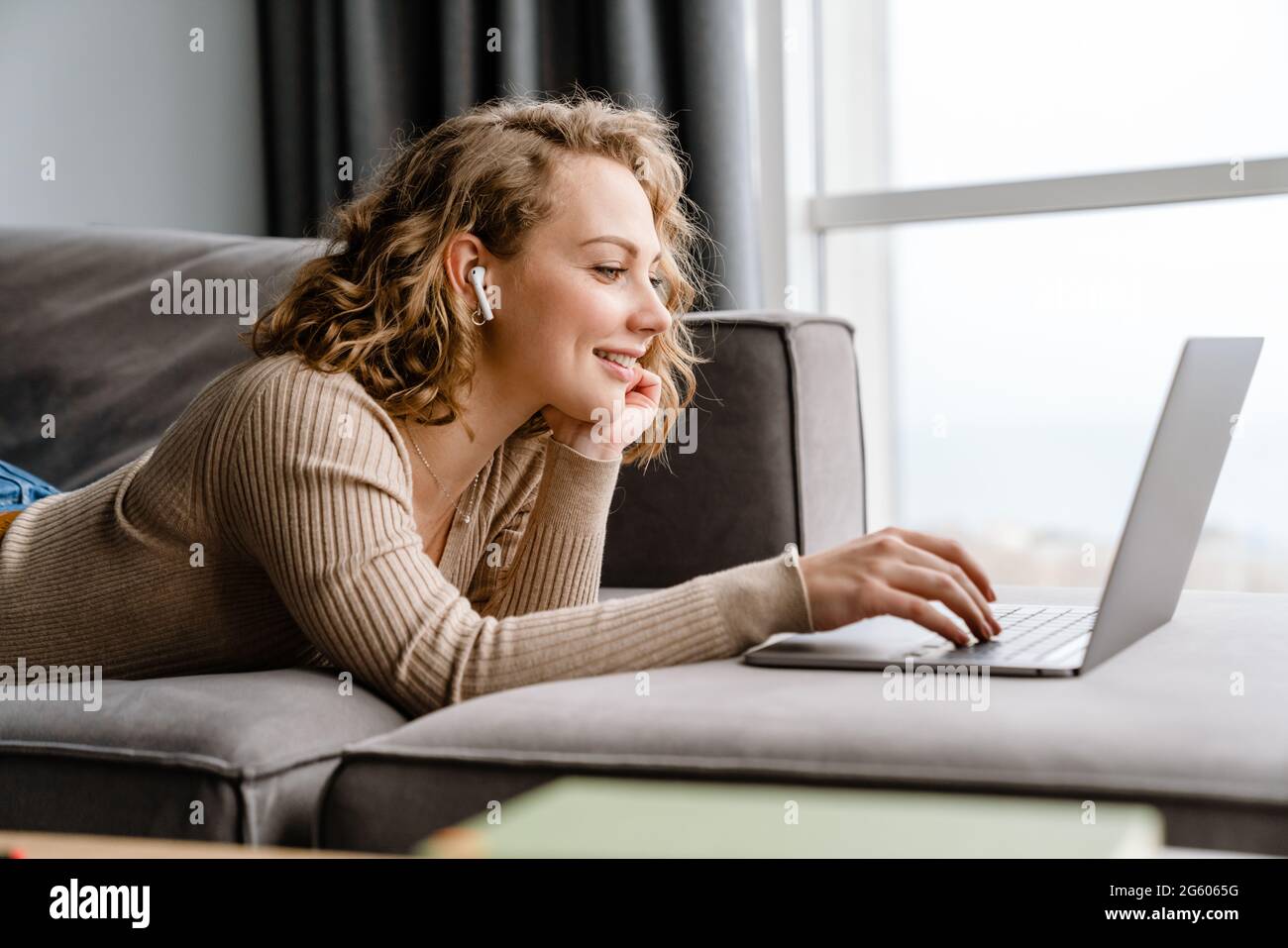 Young woman in earphones using laptop while lying on couch at home ...