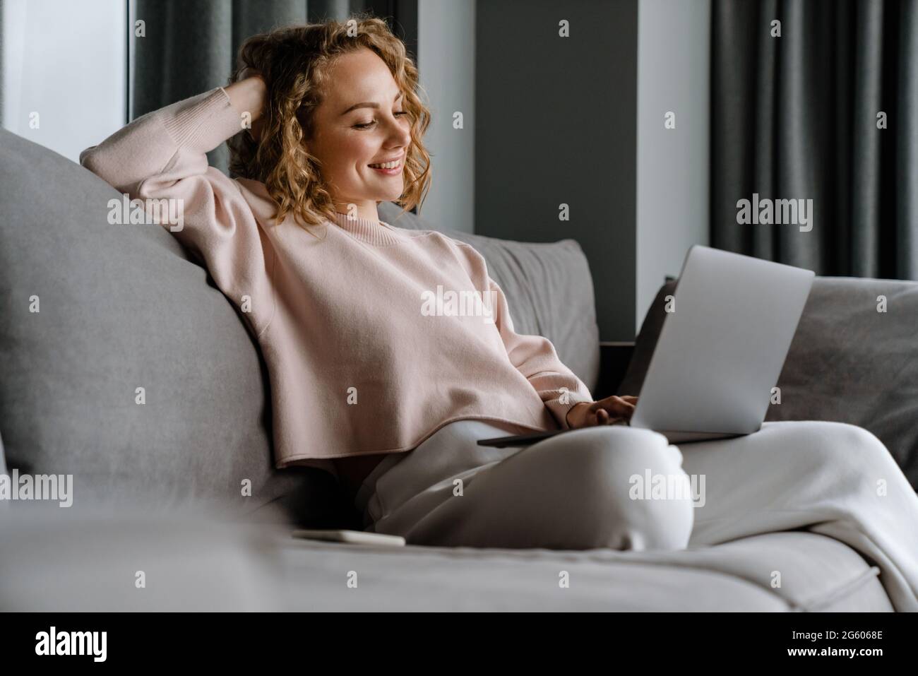 Young white woman smiling and using laptop while sitting on couch at ...