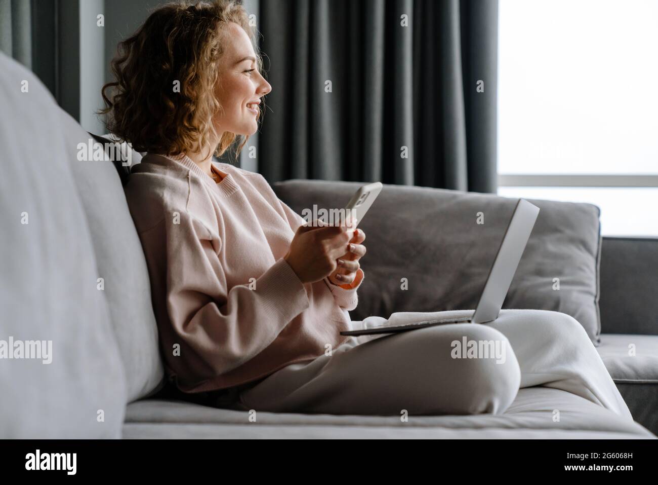 Young white woman using laptop and cellphone while sitting on couch at ...
