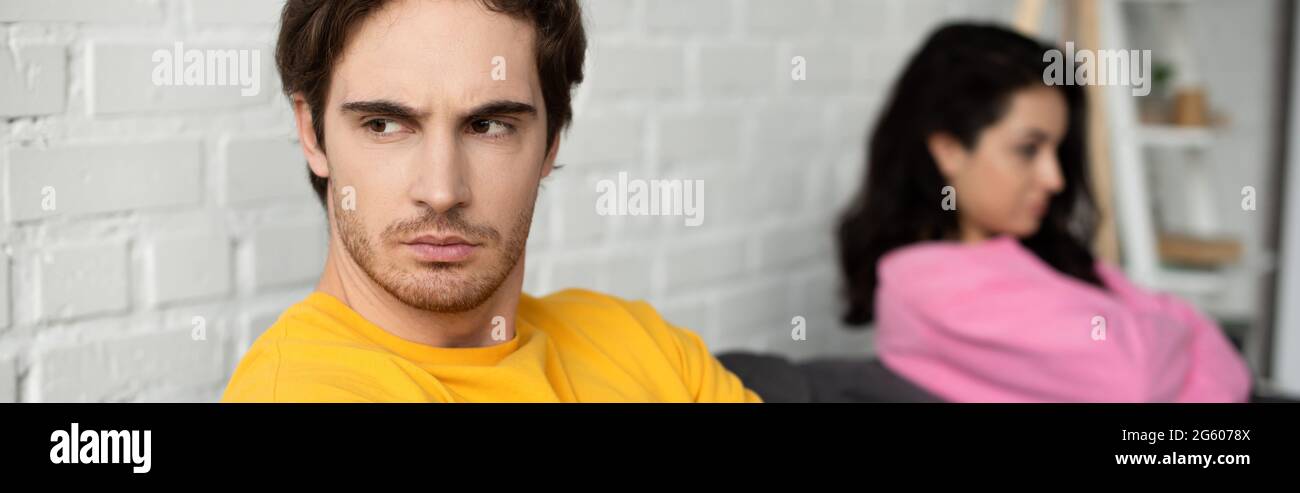 disappointed young man sitting on couch near blurred girlfriend at home ...