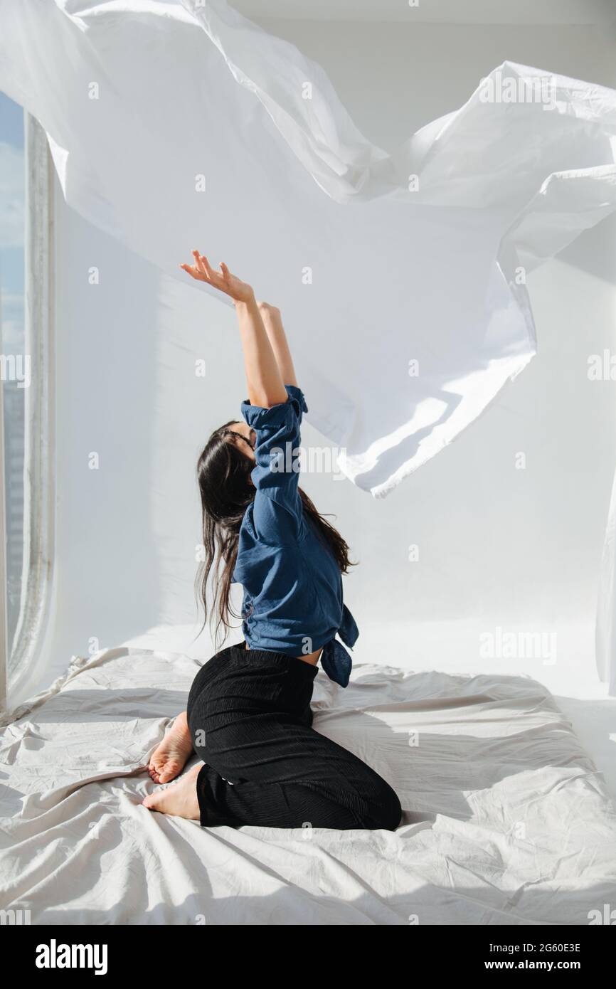 side view of young armenian woman throwing up white sheet in bedroom ...