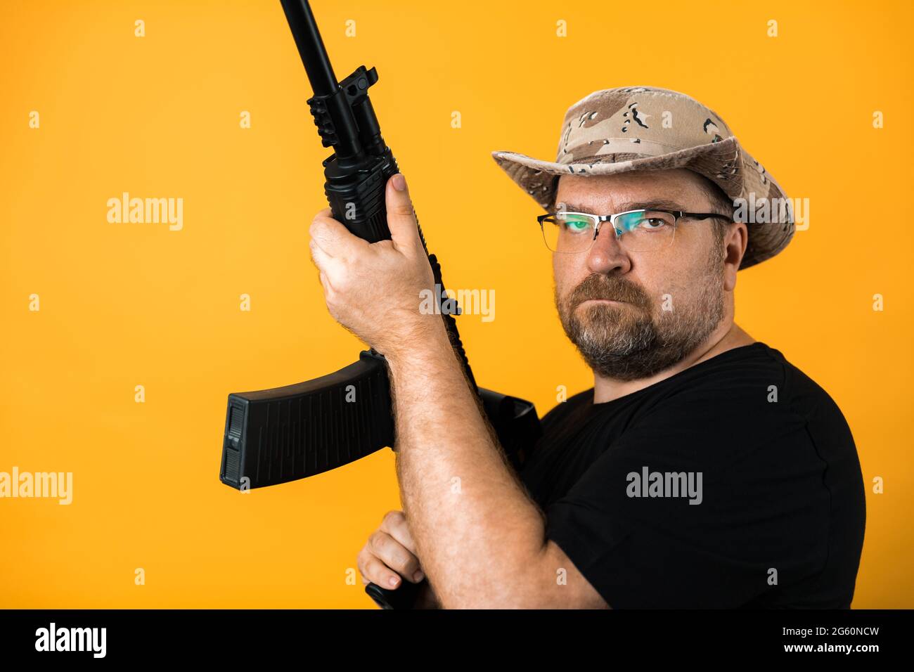 Middle aged man with rifle in black t-shirt and hat against yellow ...