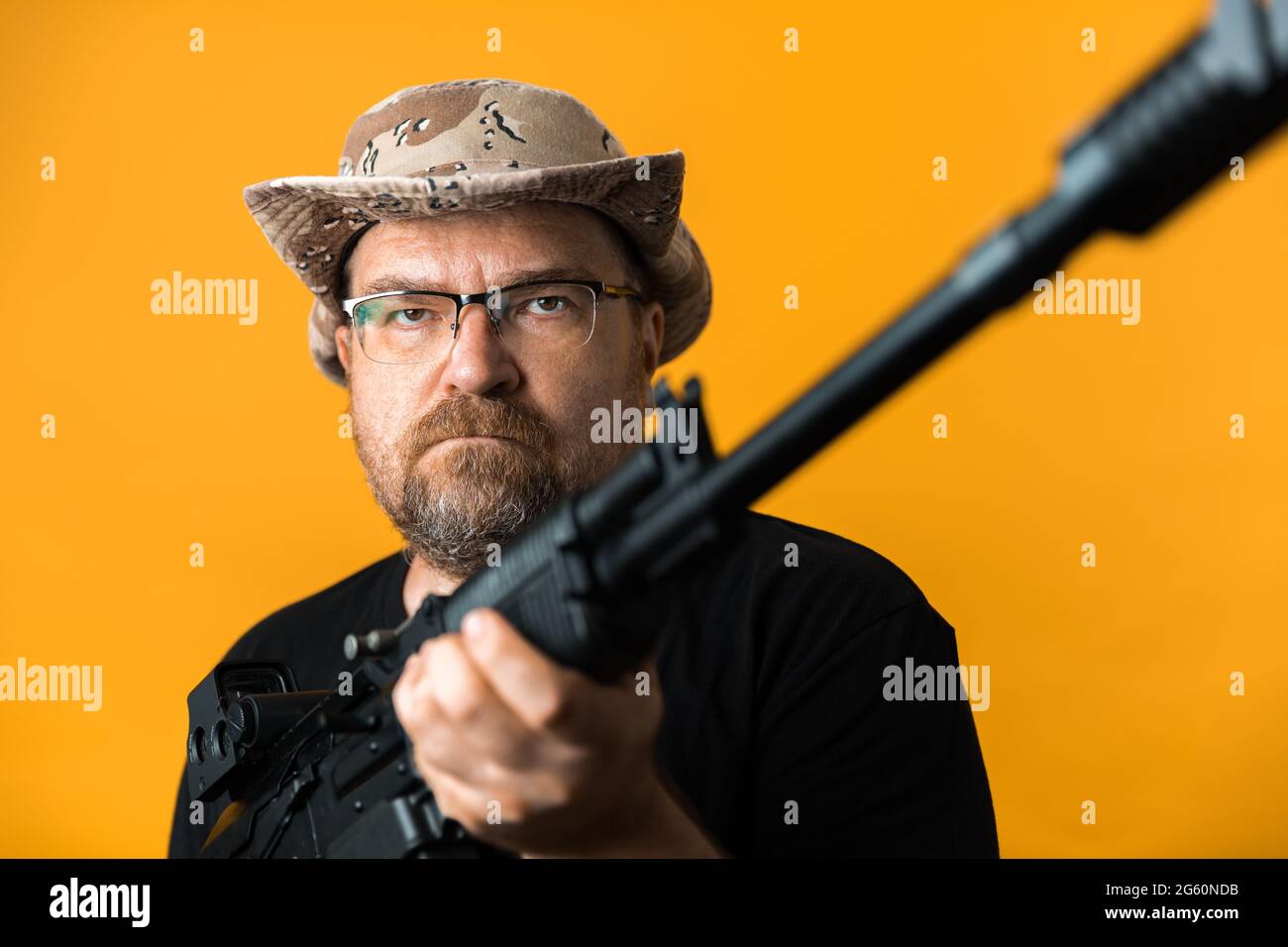 Middle aged man with rifle in black t-shirt and hat against yellow ...