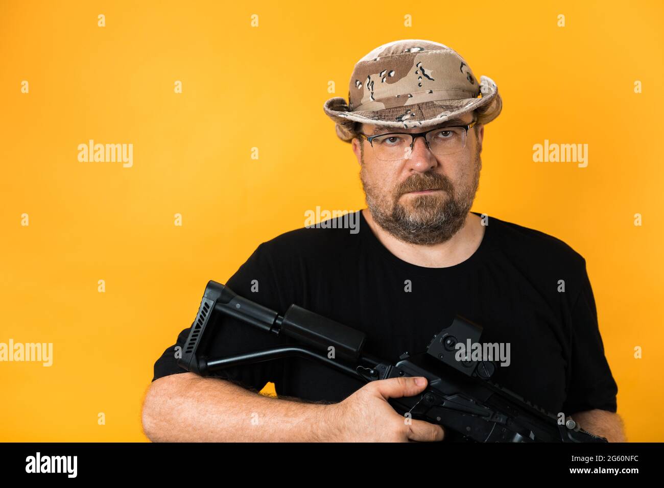 Middle aged man with rifle in black t-shirt and hat against yellow ...