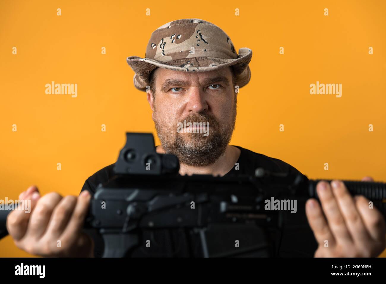Middle aged man with rifle in black t-shirt and hat against yellow ...