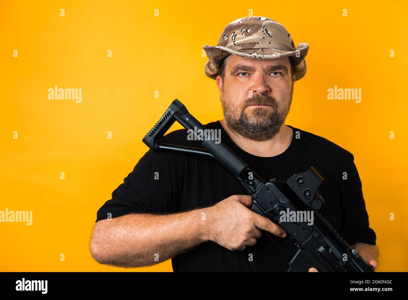 Middle aged man with rifle in black t-shirt and hat against yellow ...