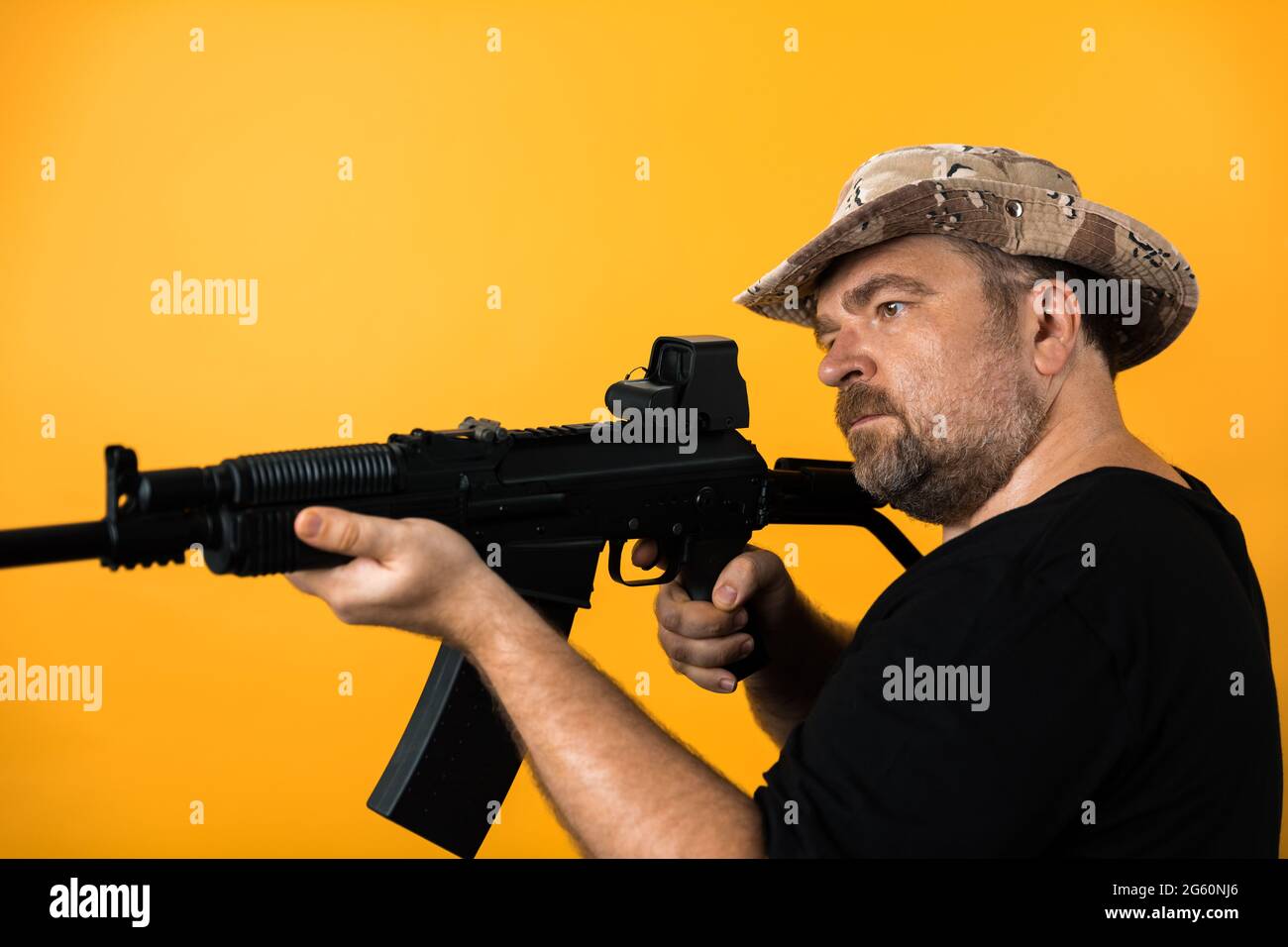 Middle aged man with rifle in black t-shirt and hat against yellow ...