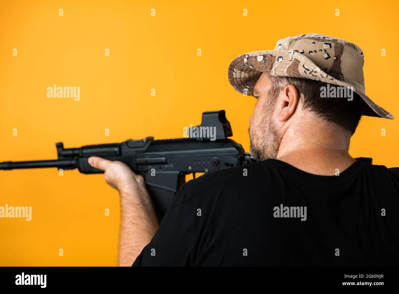 Middle aged man with rifle in black t-shirt and hat against yellow ...